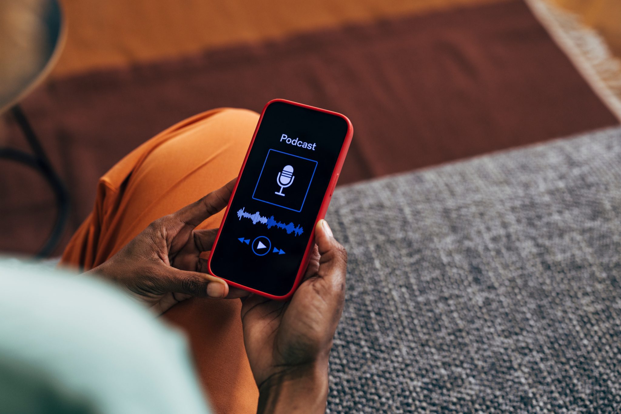 Person Holding Red Smartphone Displaying Podcast Screen Indoors — RISMedia
